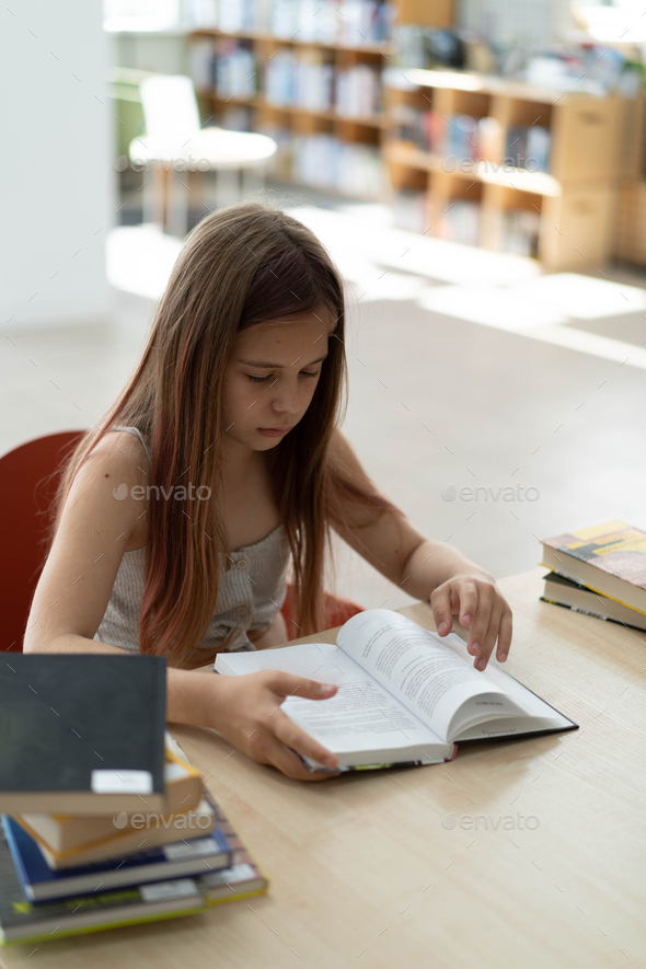 Caucasian girl in the library among stacks of books reading a book ...