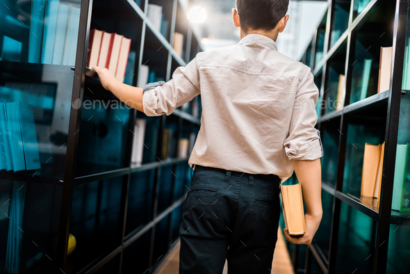 back view of boy holding book and walking between bookshelves in ...