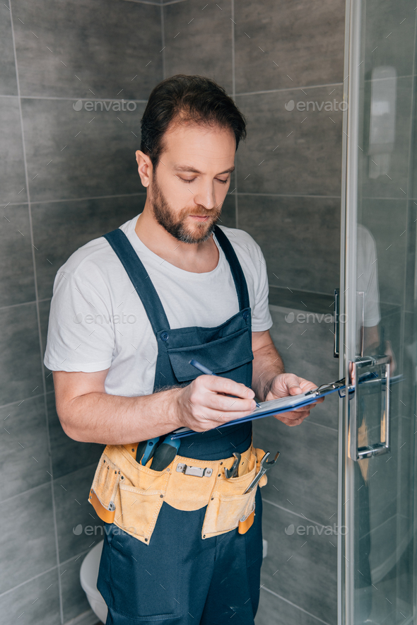 bearded male plumber making notes in clipboard while checking shower in ...