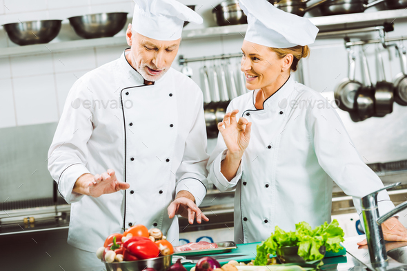 female and male chefs in uniform and hats talking while cooking in ...