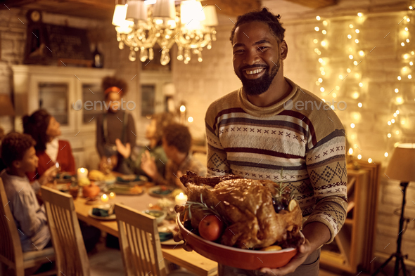 Happy black father carrying roast turkey while serving it to his family ...