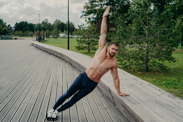 Athletic man stands in side plank pose, raises one arm, poses with ...