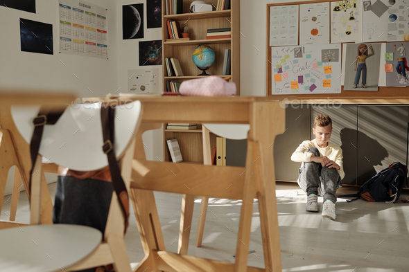 Lonely and unhappy schoolboy sitting on the floor of empty classroom ...