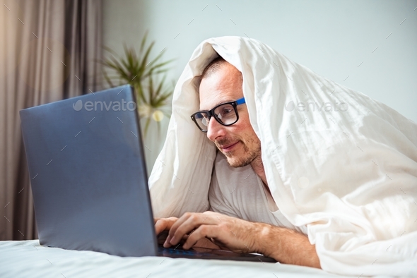 Handsome man typing a text on laptop keyboard lying under the blanket ...