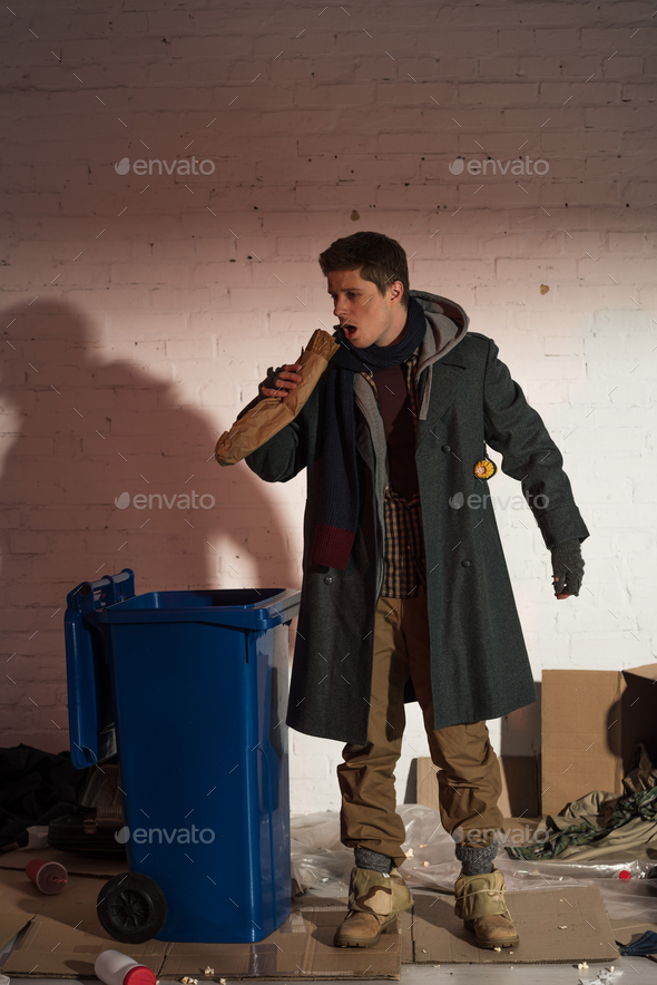 hungry homeless man eating bread baguette while standing by trash ...