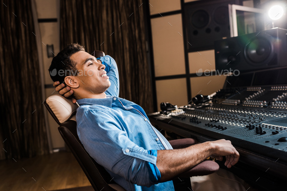 smiling mixed race sound producer relaxing in office chair in recording ...