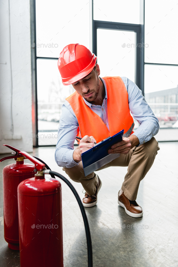 serious fireman in helmet writing on clipboard while checking ...
