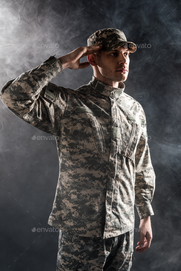 soldier in military uniform and cap giving salute on black with smoke ...