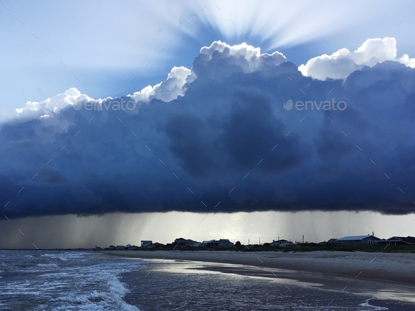 Dramatic huge cumulonimbus cloud with heavy rain pouring over sandy empty beach and sun rays on ...