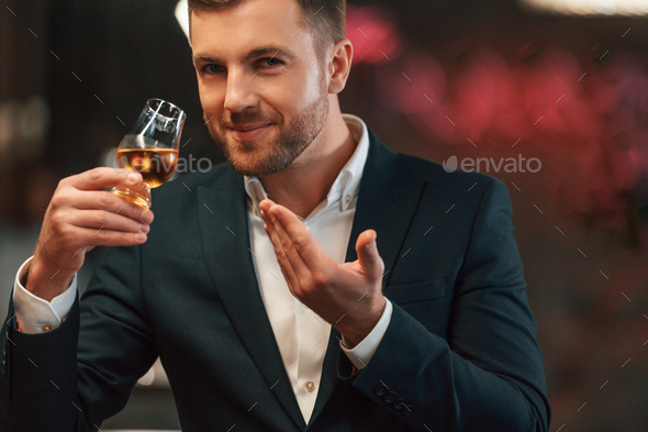 Positive elegant man in black suit is in the bar, tasting the alcohol ...