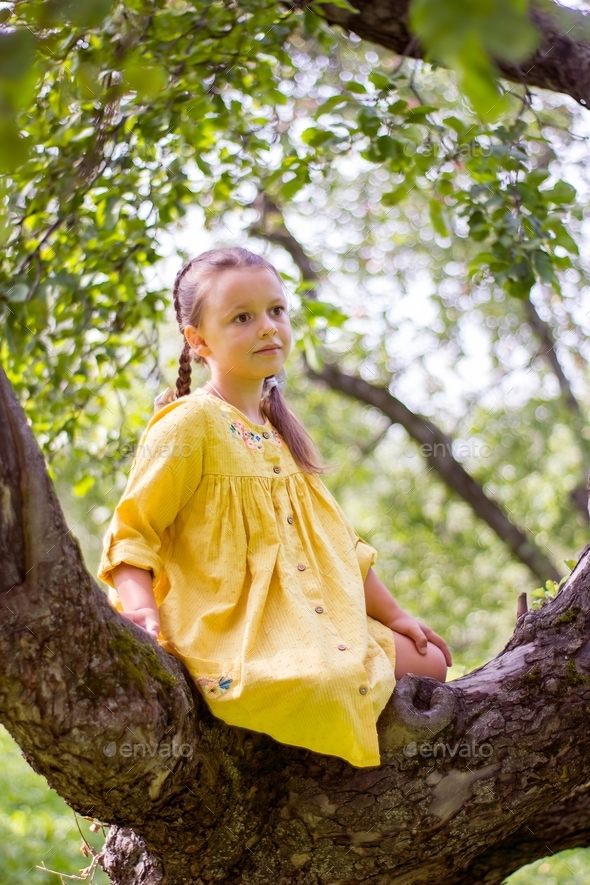 Cute little girl in a yellow dress sits on a tree branch in the garden ...