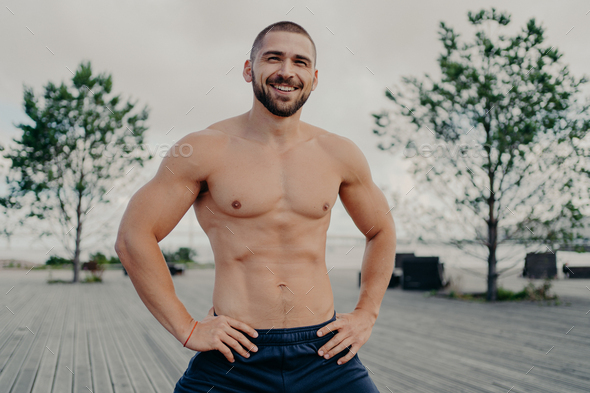 Happy handsome man with thick bristle keeps hands on waist, stands with ...