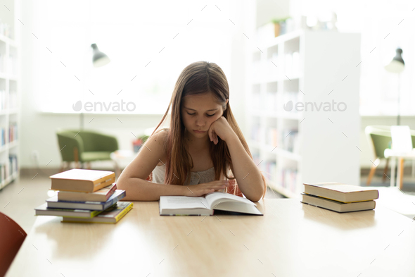 Caucasian girl in the library among stacks of books reading a book ...
