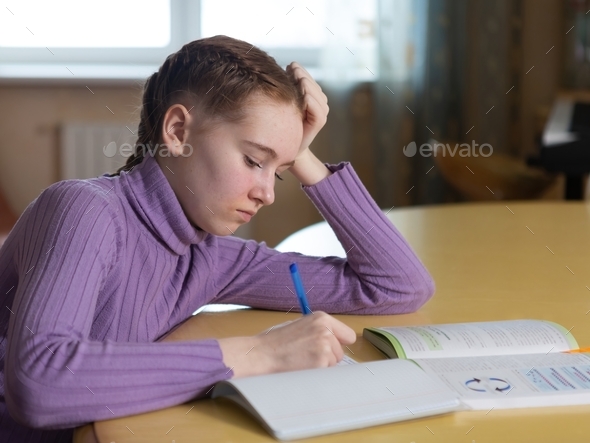 Teenage girl doing homework. She sits at the table at home and writes ...