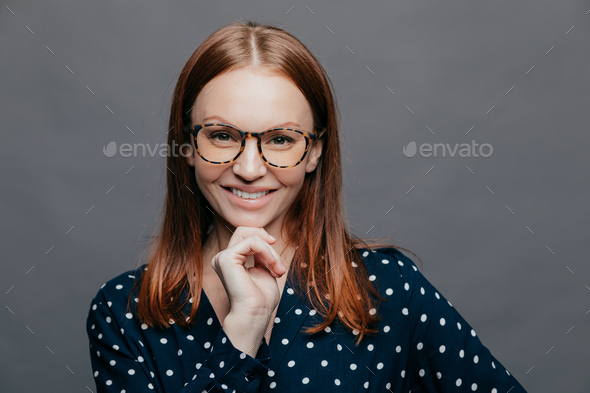 Happy delighted young woman keeps one hand under chin, smiles happily ...