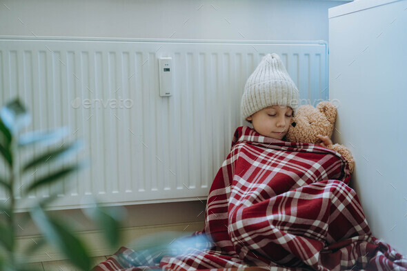 cute little boy wrapped id plaid wearing knit hat sitting by heater ...