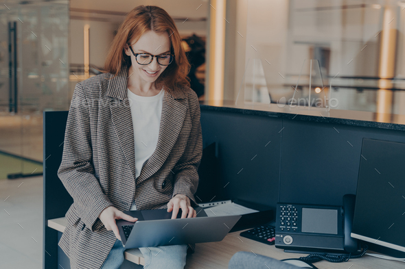 Beautiful redhead businesswoman sitting on top of desk and using laptop ...