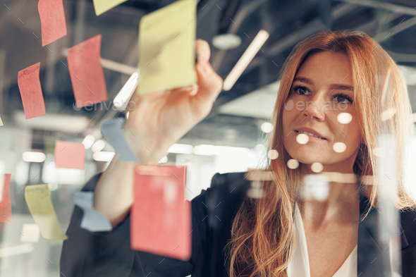 Businesswoman writing on sticky notes on glass wall while working in ...