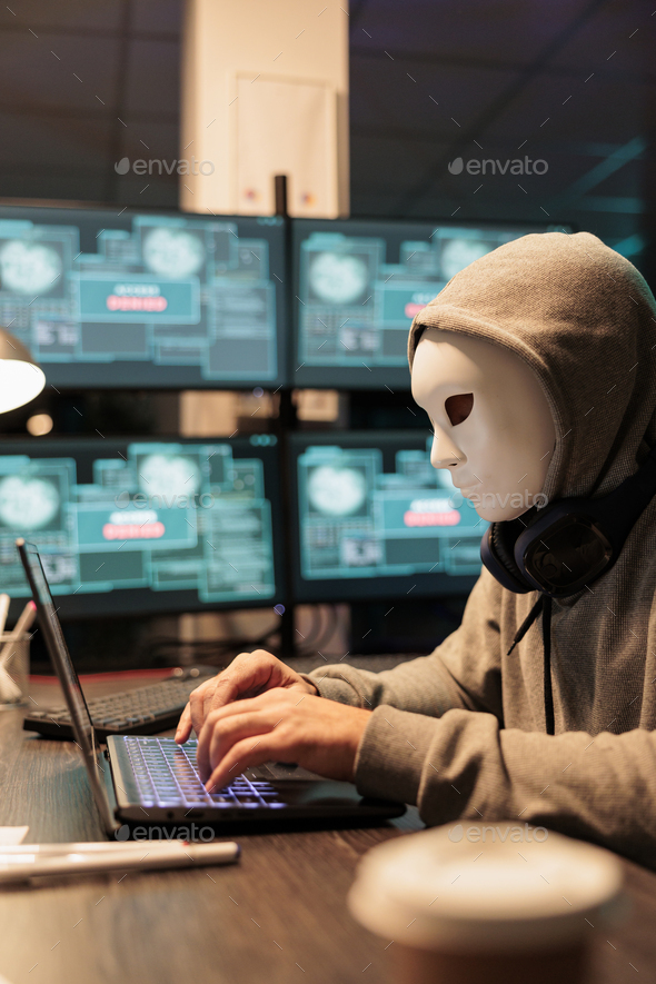Scary male scammer wearing mask hacking database Stock Photo by DC_Studio