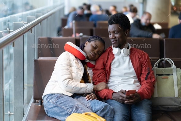 African couple of tourists in waiting area of airport terminal sit, nap ...