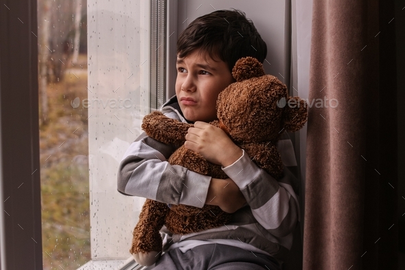 Sad boy looks out the window hugging a teddy bear. Bad weather mood ...