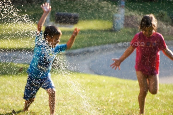 Two children running through a sprinkler and playing outdoors in the ...