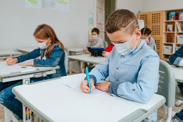 Elementary school students in face masks sitting at the desks in the ...