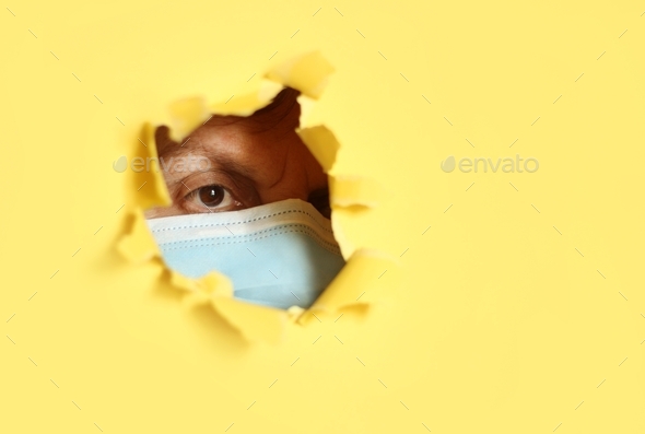 Female face with a face mask peeking out of a hole in yellow paper ...