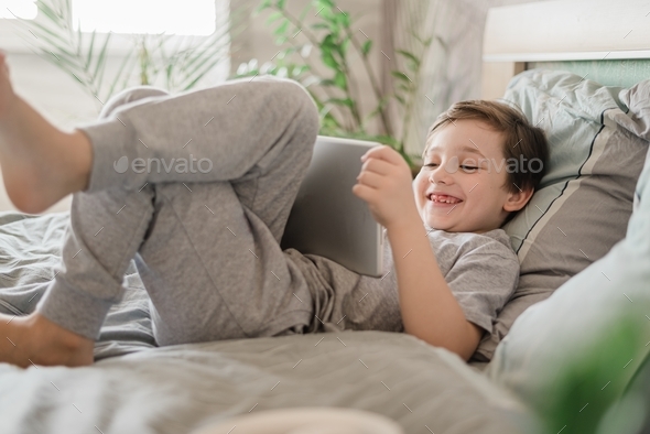 Little kid boy using tech and lying on bed in the light and airy room ...