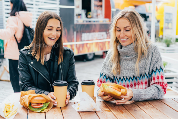 Two satisfied happy hungry women eat burgers in the street market ...