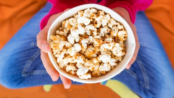 Hands hold a bucket of popcorn while watching a movie at home. Top view ...