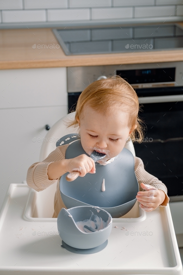 Self feeding of a grubby toddler sitting in a highchair, wearing bib ...