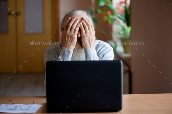 a lonely old man with a gray beard hides his face in his palms sitting ...