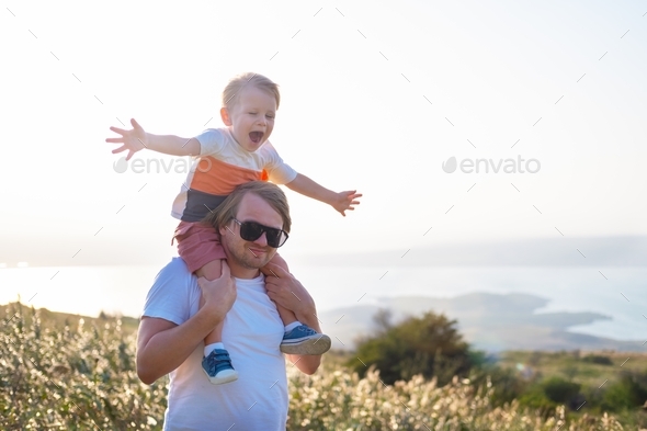 smiling kid blond boy sitting on dad shoulders in nature on a windy and ...
