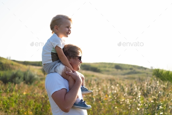 smiling kid blond boy sitting on dad shoulders in nature on a windy and ...