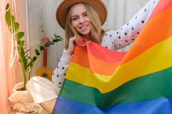 Young blonde millennial hippie woman with rainbow LGBTQ flag at home ...
