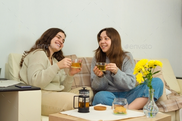 Cheerful young women are sitting on the sofa drinking tea and talking ...