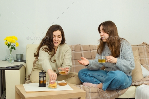 Young women sit on the sofa and communicate, pass cookies to each other and drink tea. Stock ...