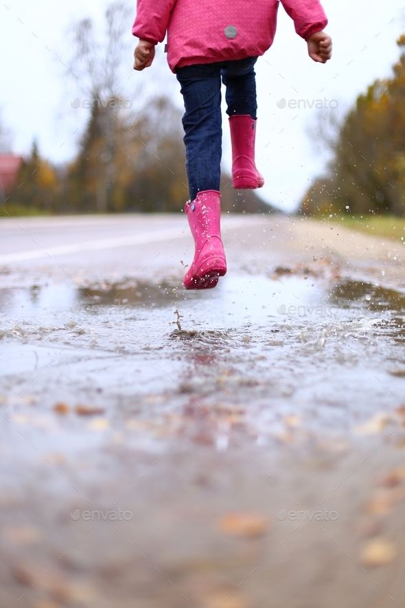 Little girl in a pink jacket and pink rubber boots jumps through puddles along the road Stock ...