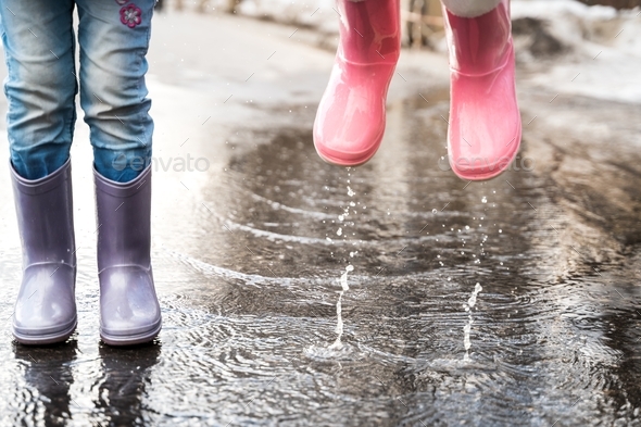 Little girls in pink, purple waterproof rubber boots cheerfully jumps through puddles on street ...