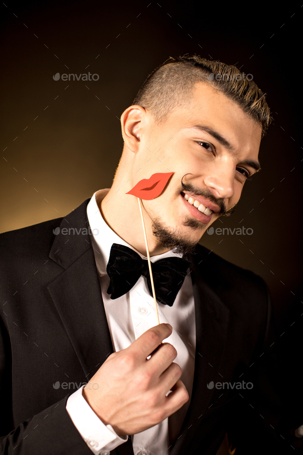 Smiling young man in tuxedo posing with red lips on stick Stock Photo ...