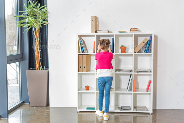 Back view of cute little girl choosing book on bookshelves Stock Photo ...