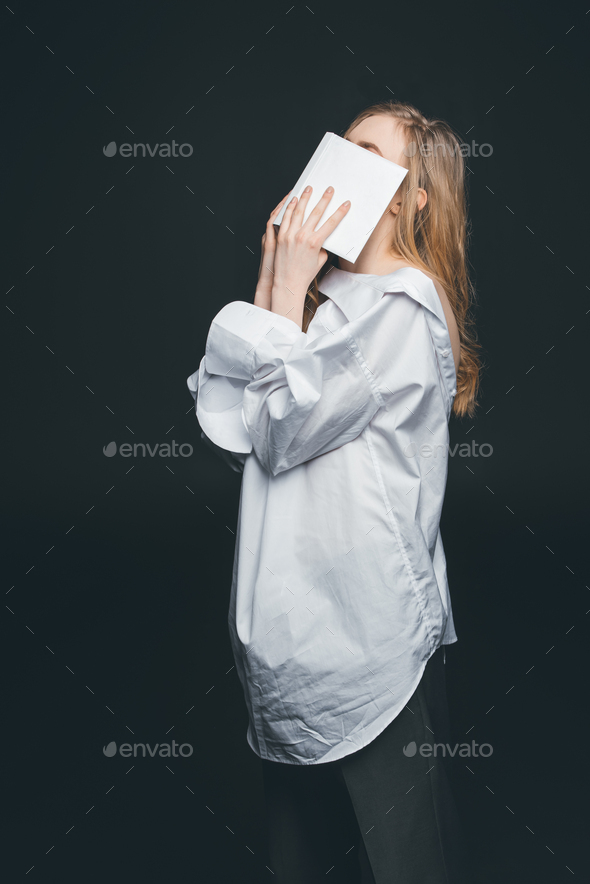 Cute blonde fashion girl hiding face by book, posing in studio on black ...