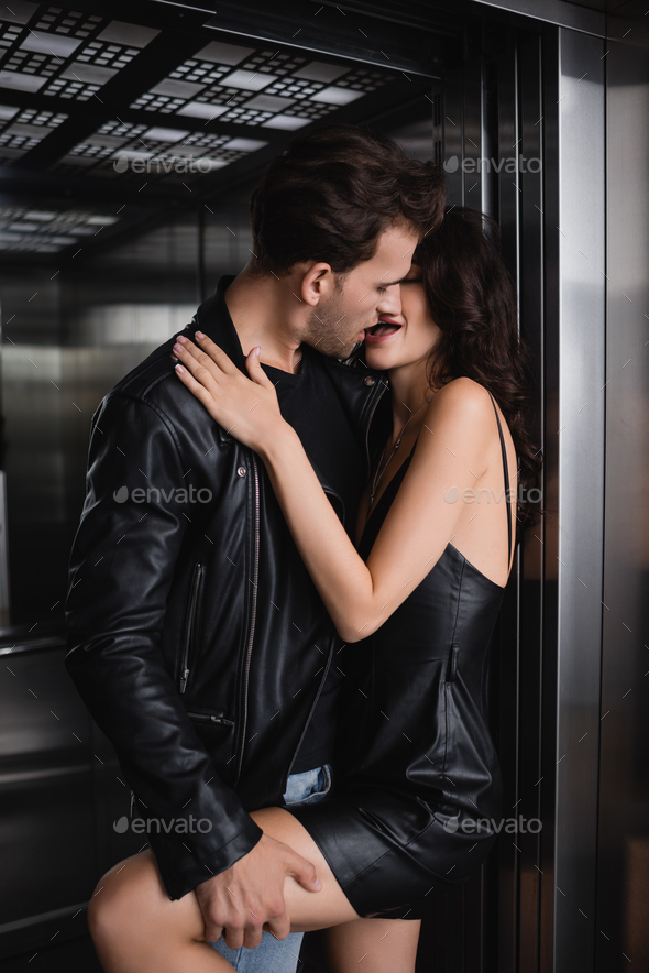 Man in leather jacket kissing and holding leg of seductive woman in black dress in elevator ...