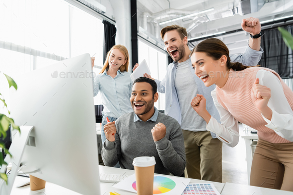 Cheerful multiethnic businesspeople showing yeah gesture near computer ...