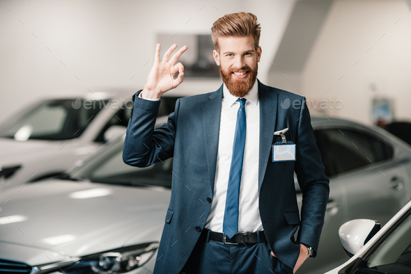 salesman in suit showing ok sign and looking at camera in dealership ...