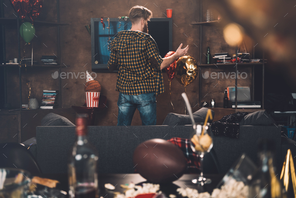 Back view of confused young man gesturing while standing in messy room ...