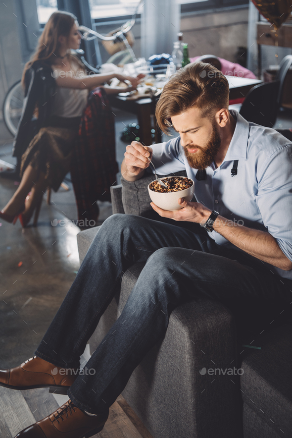 tired man eating corn flakes at home after party, woman behind Stock ...