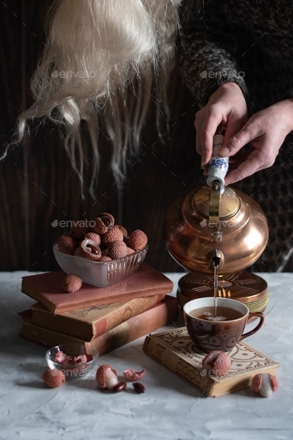 A woman pours tea from a copper teapot into a cup, lychee on stack of ...