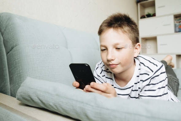 Teenage boy lying on couch using smartphone texting playing video games ...